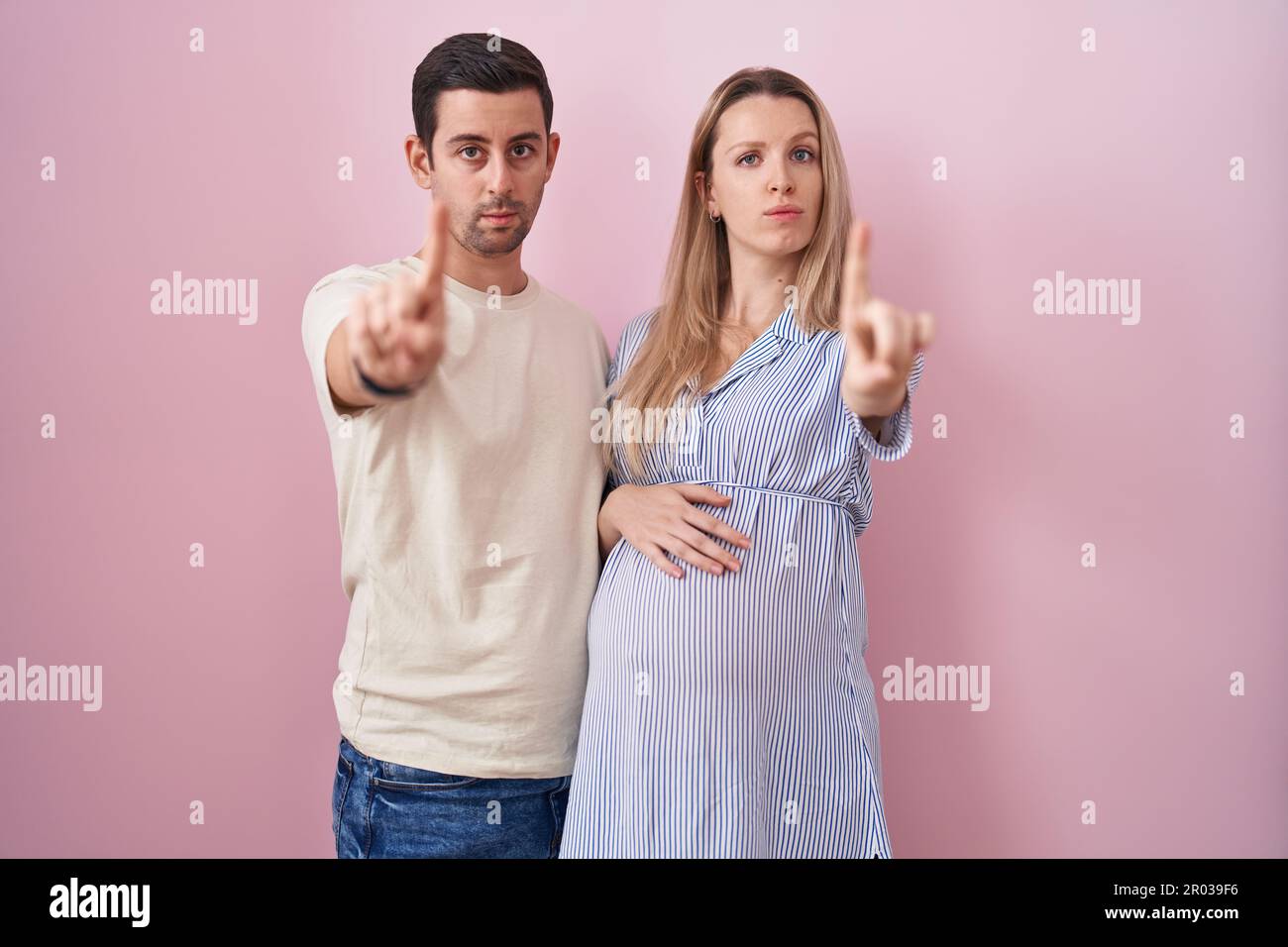 Young couple expecting a baby standing over pink background pointing ...