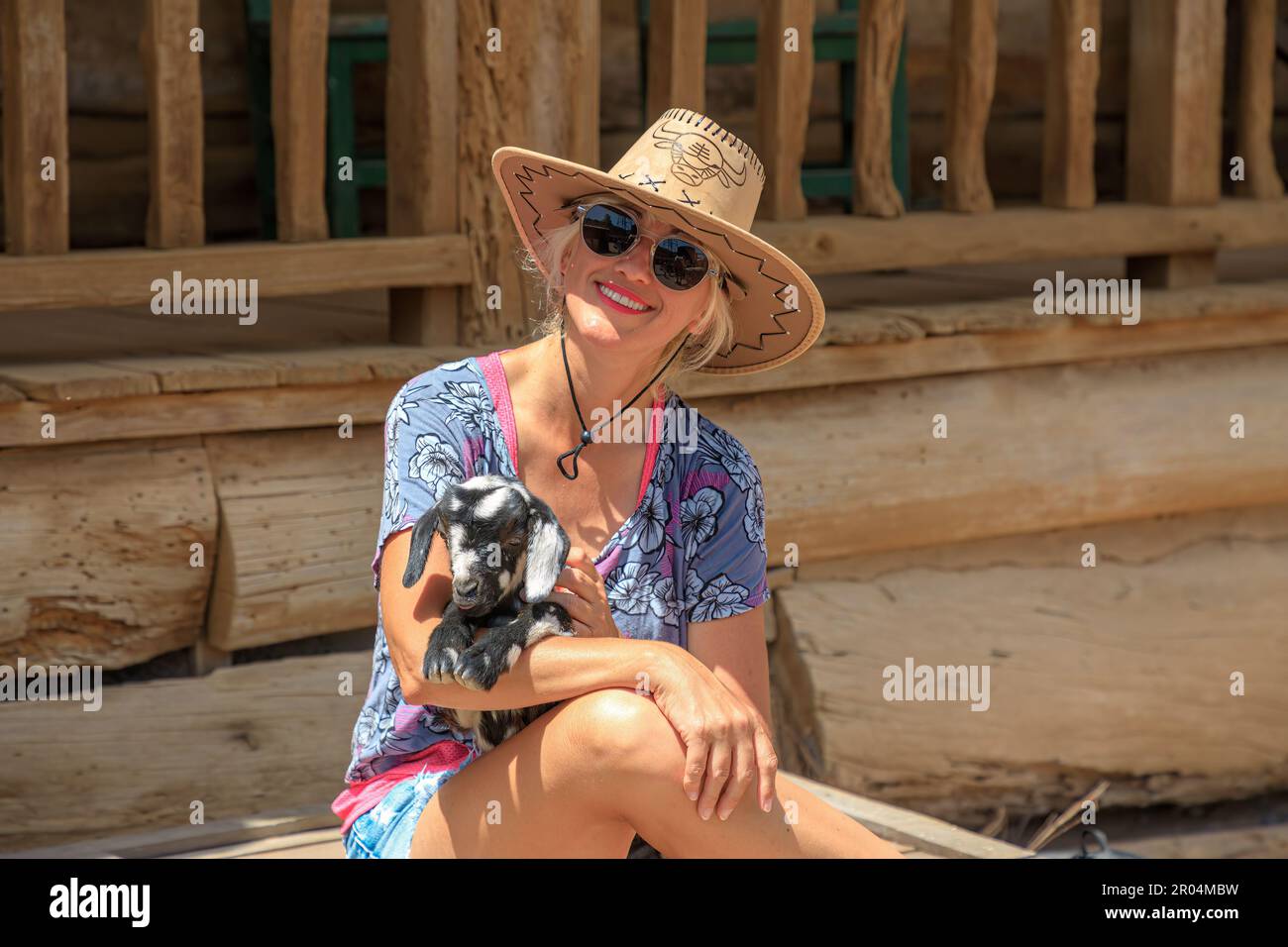 On the porch of a farm, a woman sits with a small baby goat, its tail ...