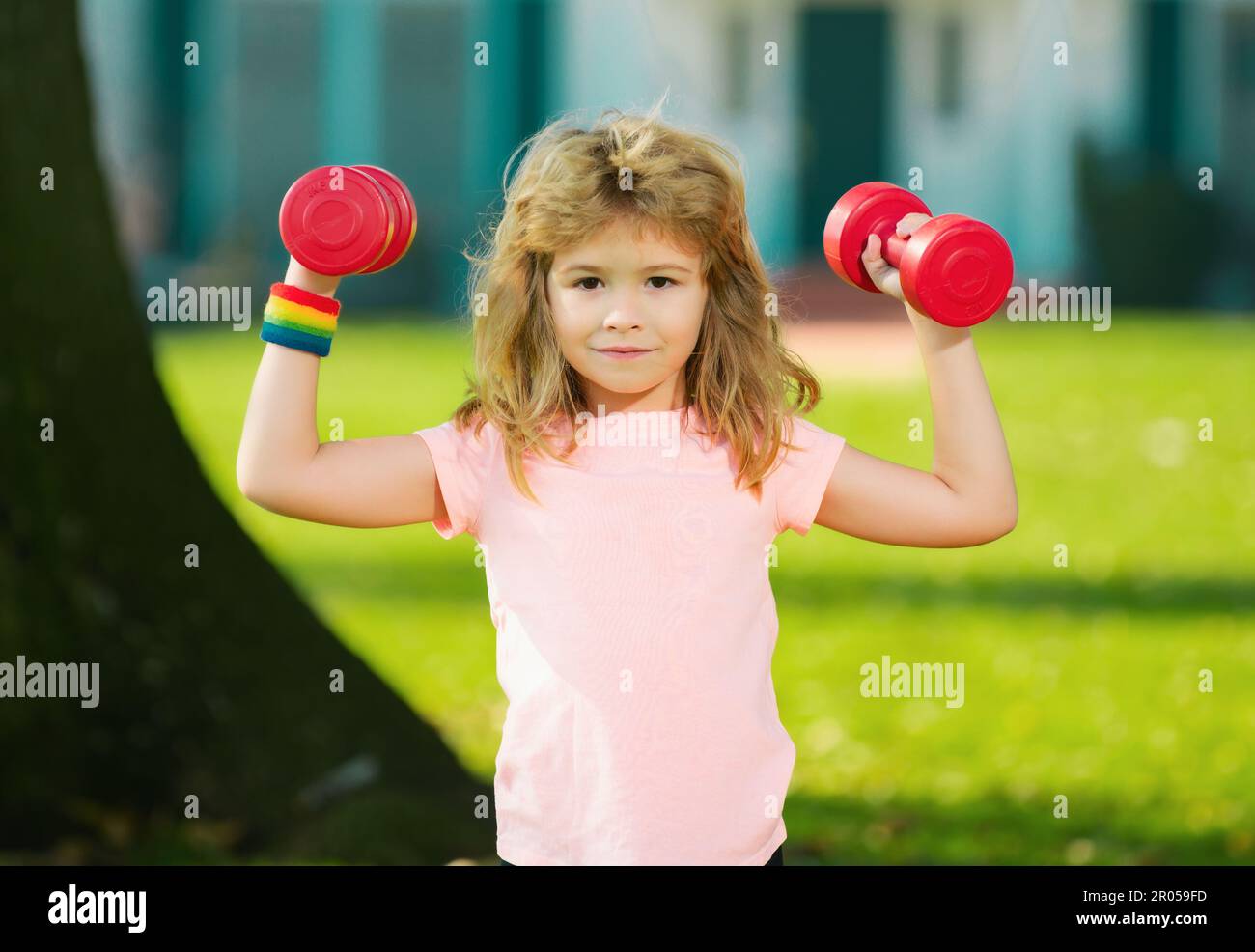 Cute child boy pumping up arm muscles with dumbbell outdoor in summer park. Fitness kids with ...