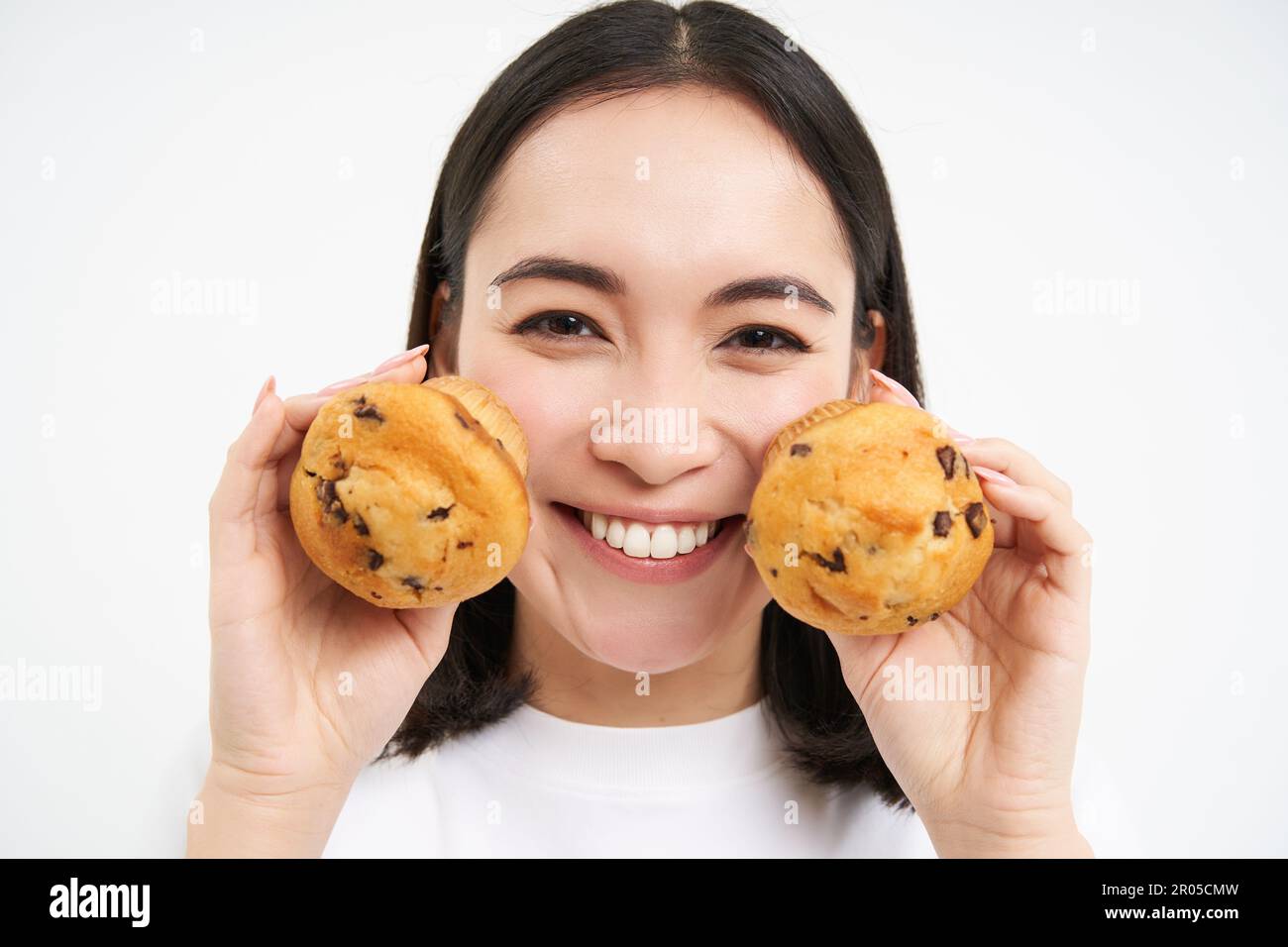 Close up portrait of asian woman, shows two cupcakes near face and ...