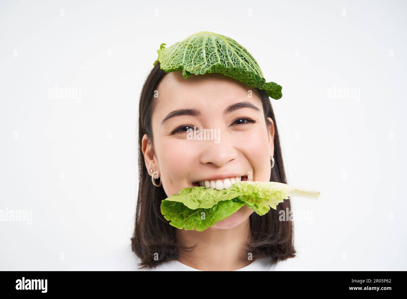 Beautiful asian girl with lettuce on head, smiling and eating cabbage ...