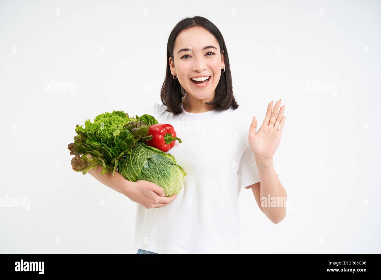 Enthusiastic asian woman, holding vegetables and waves hand, say hello ...