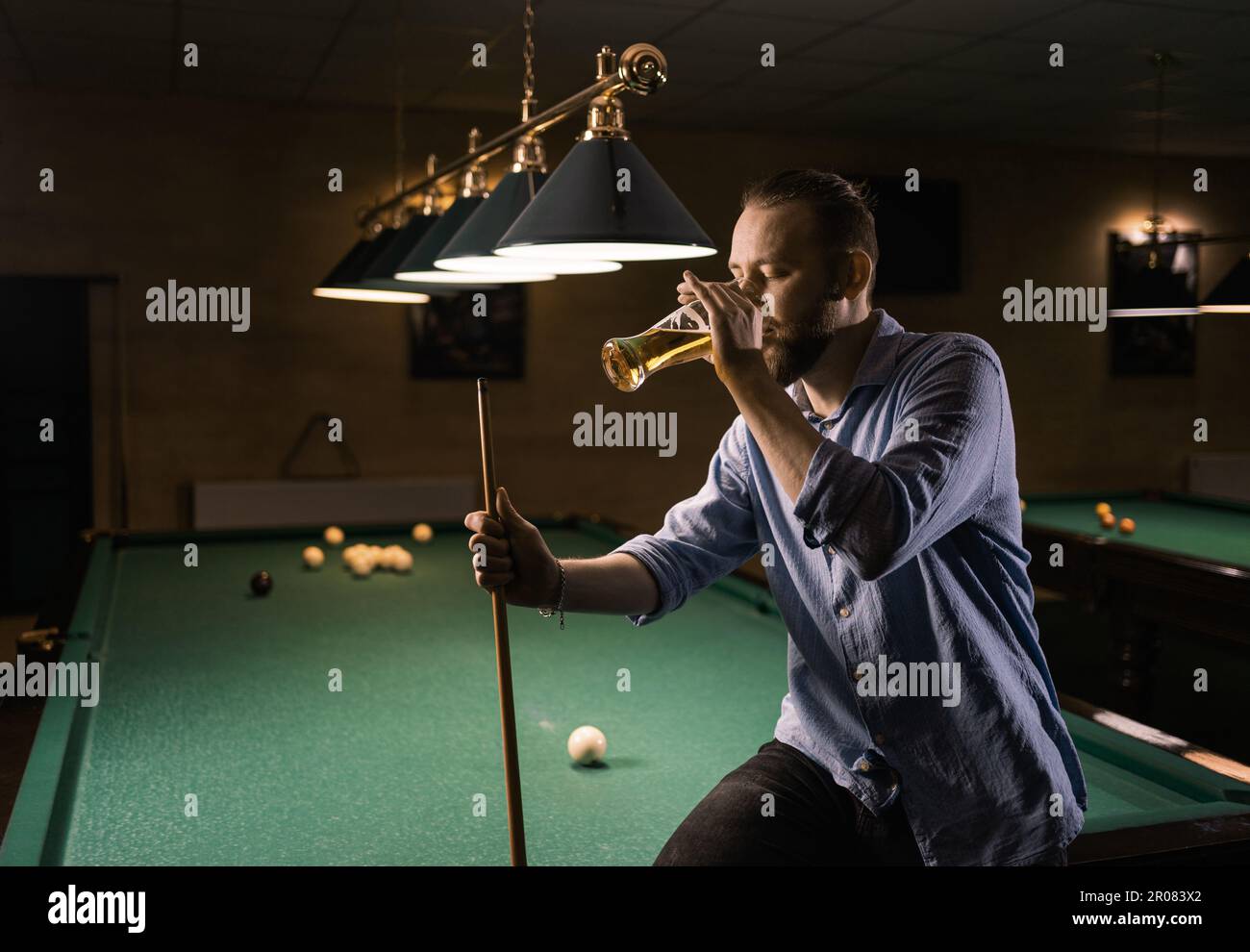 Young professional man playing billiards and drinking beer in the dark ...