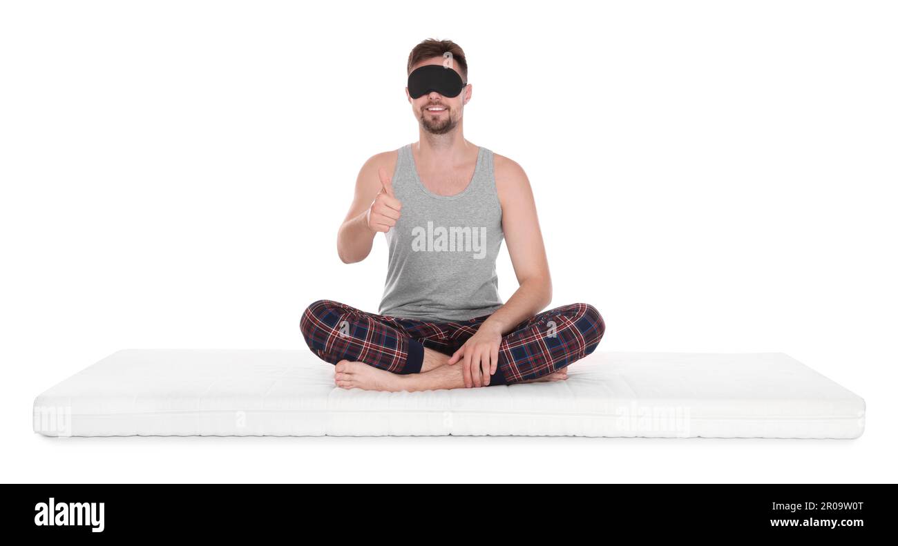 Smiling man in sleeping mask sitting on soft mattress and showing thumb ...