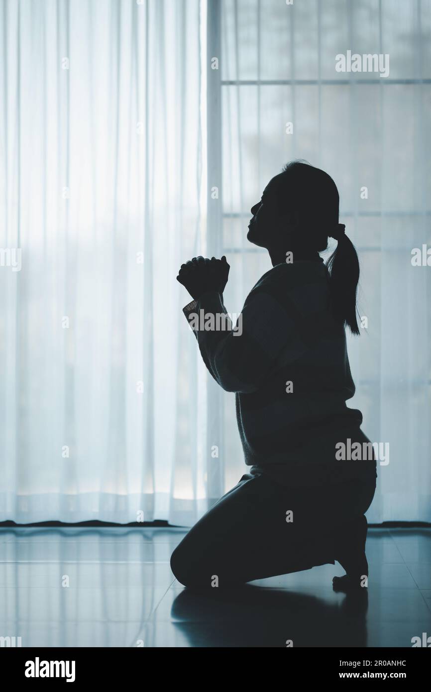 silhouette of woman kneeling and praying in modern church at sunset time. Female catholic prayer ...