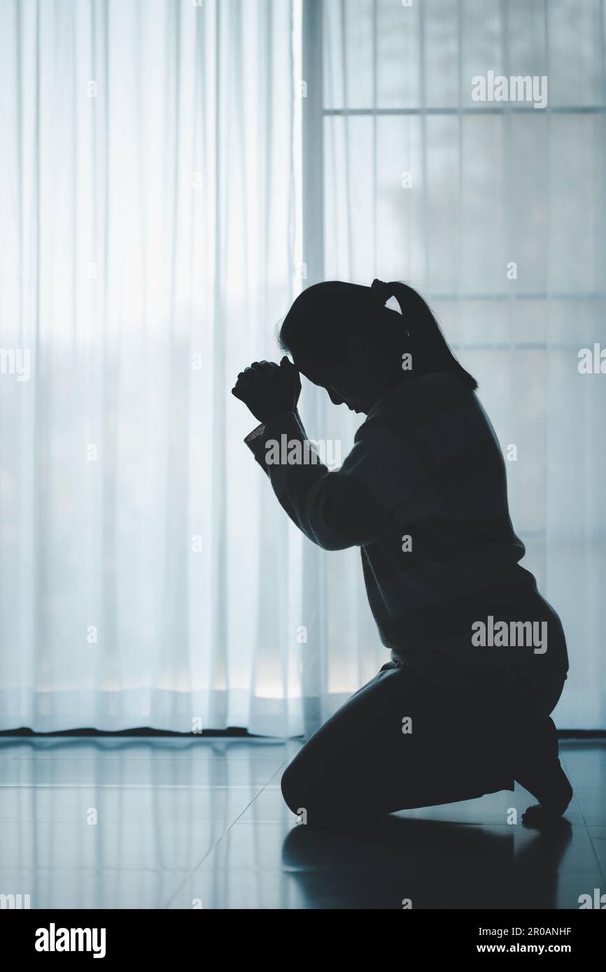 silhouette of woman kneeling and praying in modern church at sunset time. Female catholic prayer ...