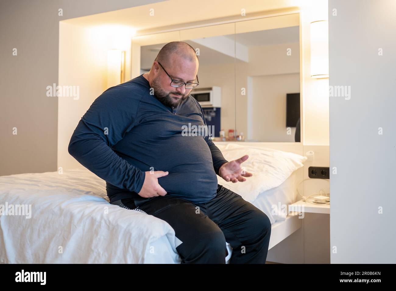 Huge man suffering from extra weight touching stomach sitting on bed in ...