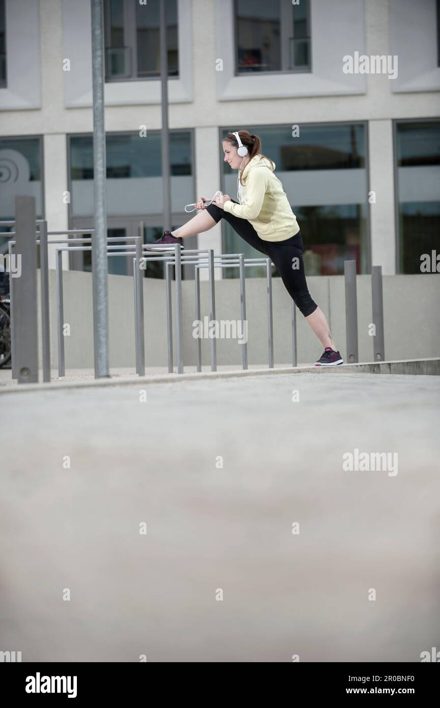 Young woman stretching her leg on railing and listening to music ...