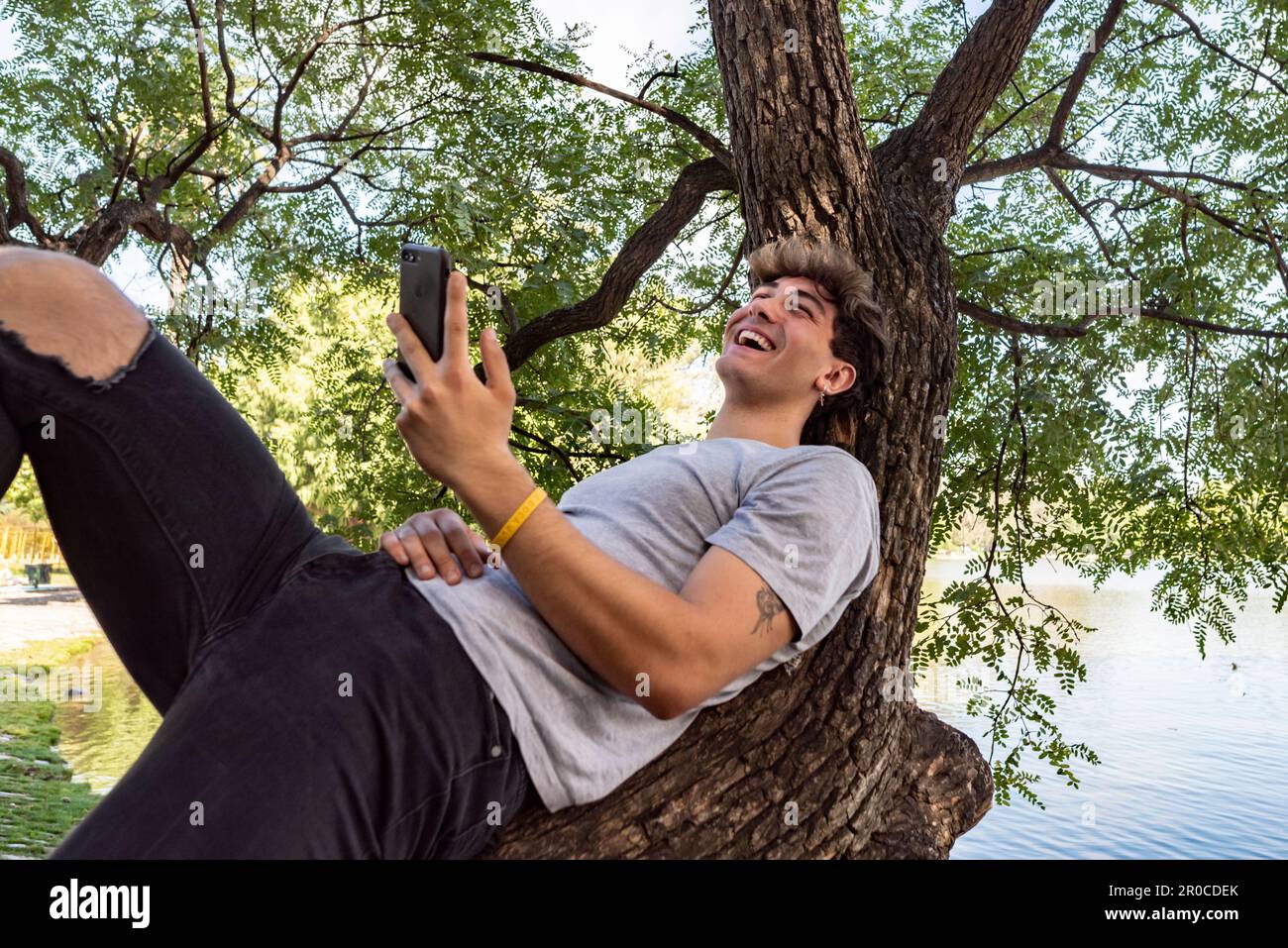 Young man leaning on a tree trunk, laughing, and using his smartphone ...
