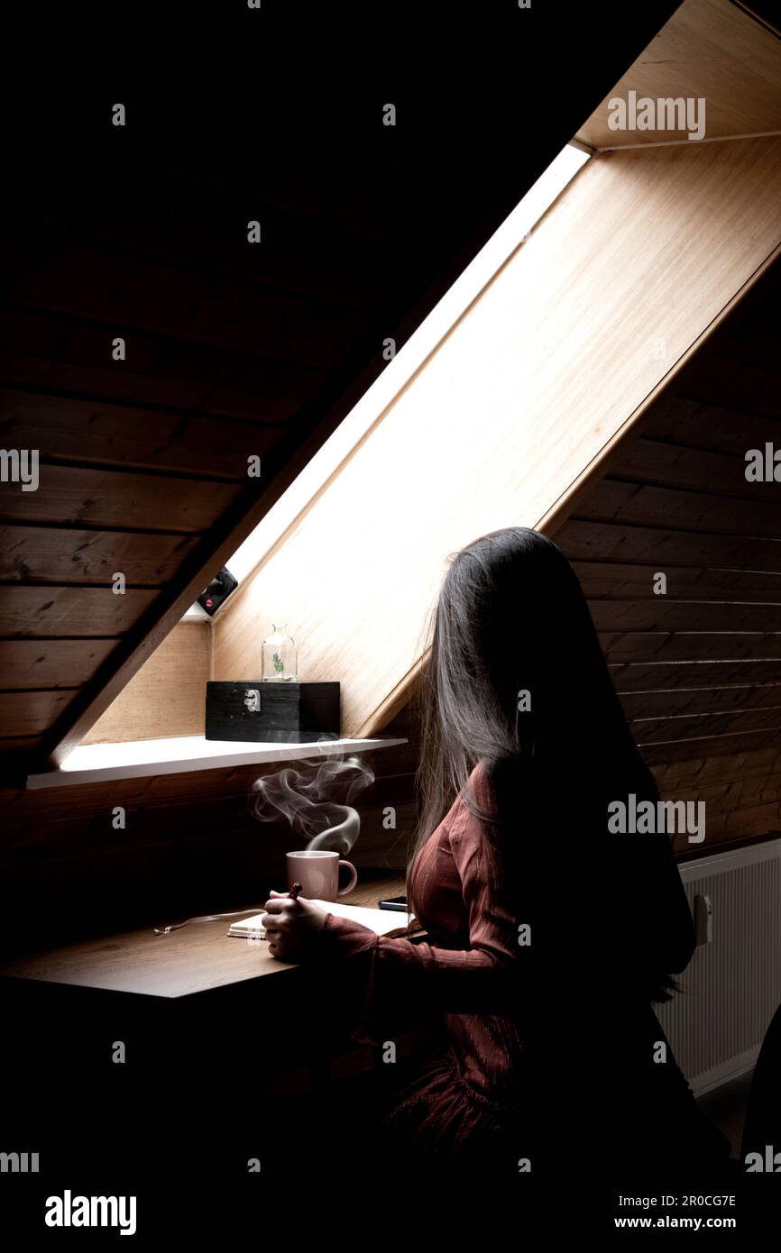 Young girl with long hair writing next to a attic window drinking tea ...