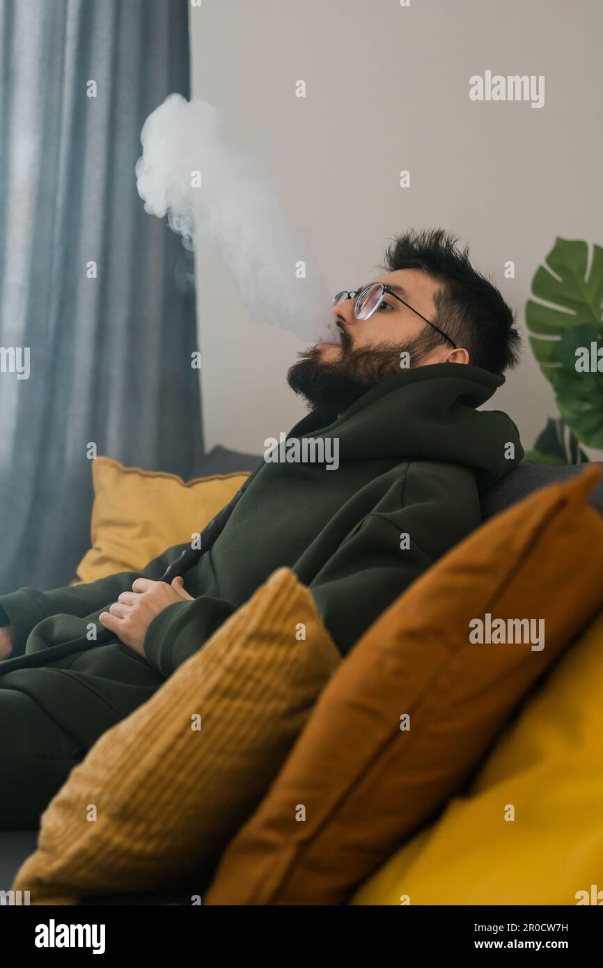 Bearded millennial or gen z man smoking hookah while relaxing on sofa ...