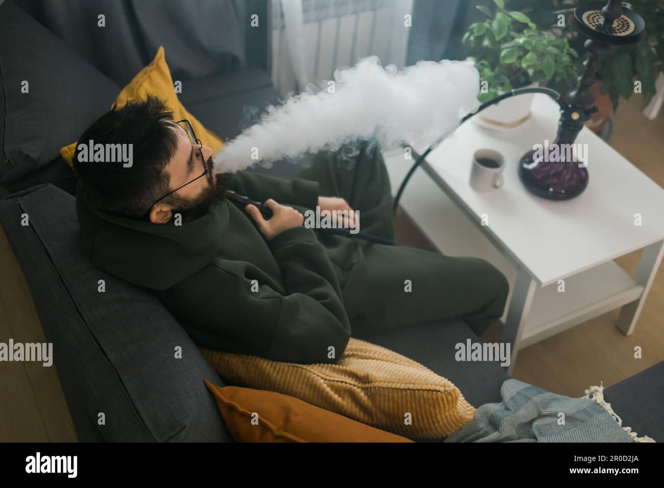 Bearded millennial or gen z man smoking hookah while relaxing on sofa ...