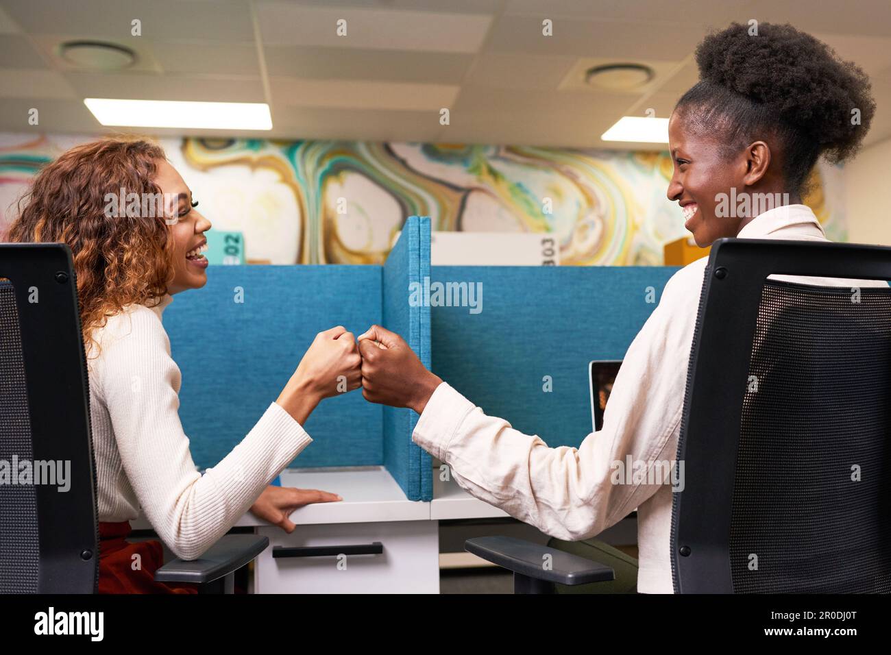 Two multi-ethnic women fist bump in modern office, across desk divider ...