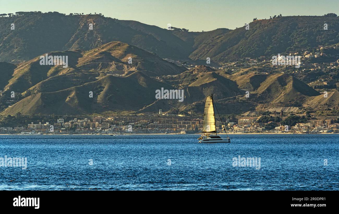 Sailboat crosses the Strait of Messina in the warm light of the sunset ...
