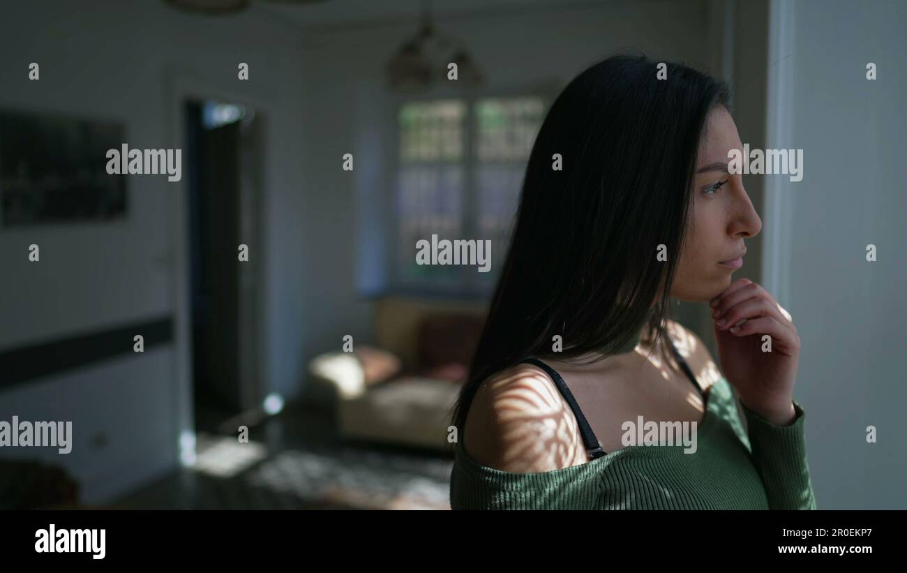 One contemplative young woman standing by window looking outside with ...