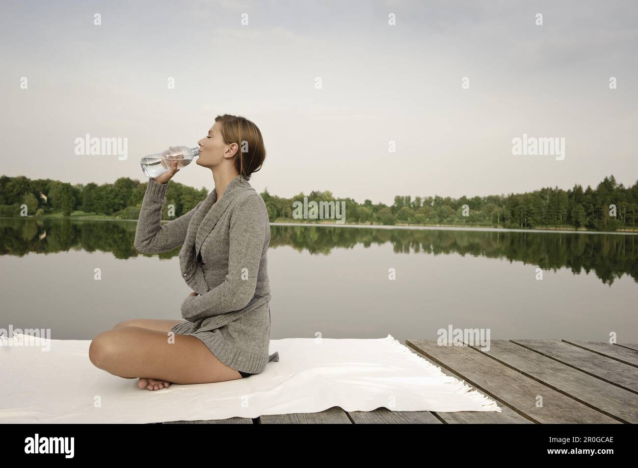 Young woman drinking a bottle of water while sitting on a jetty at lake ...