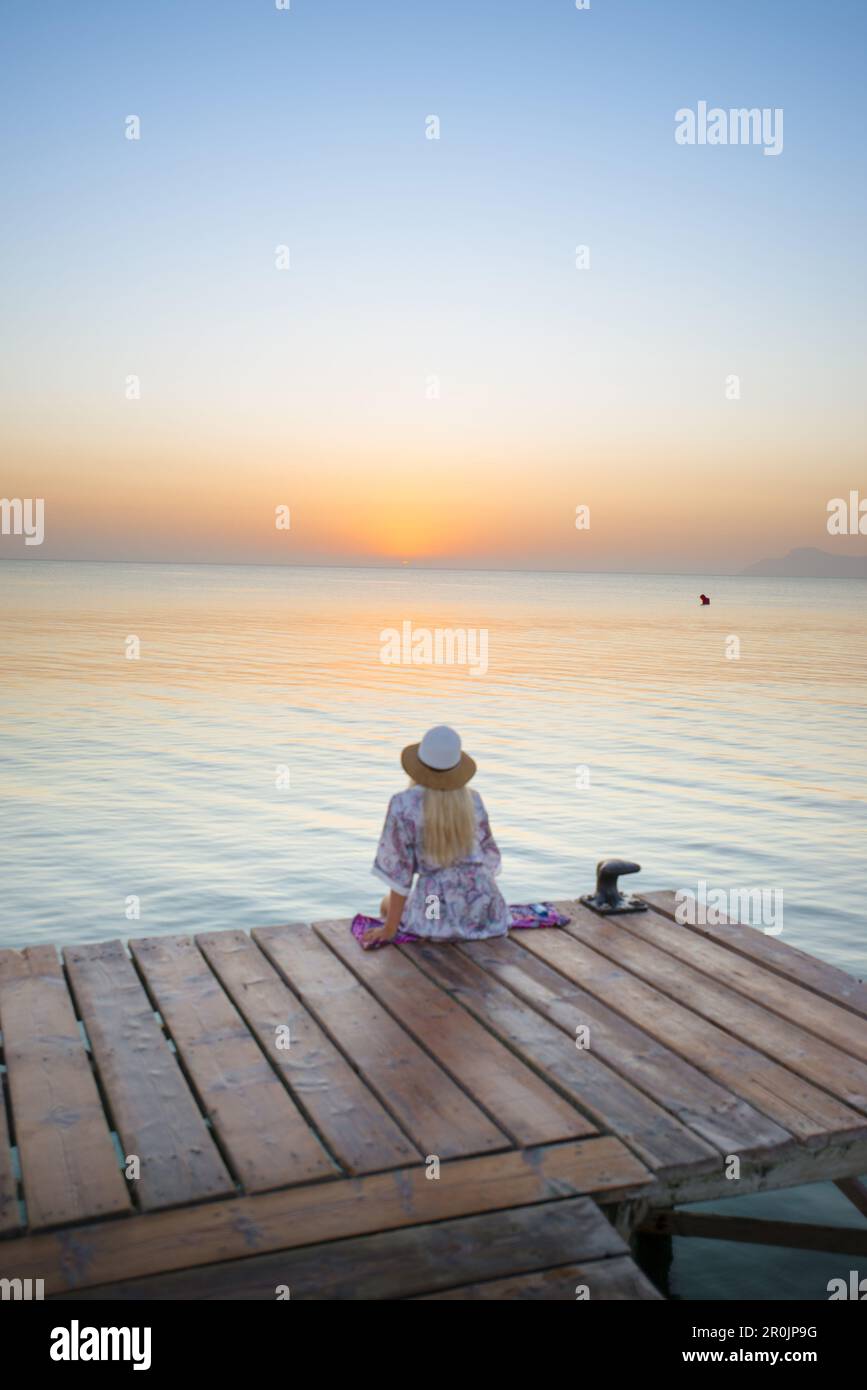 Young blond-haired woman sitting on the end of a long pier in the ...