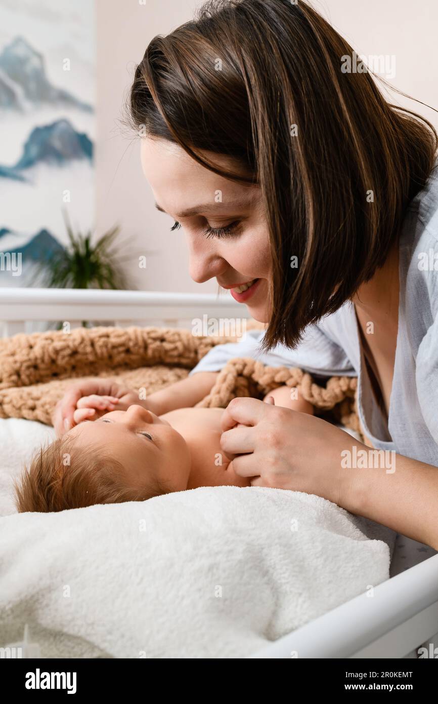 Smiling young mother hold hands of laying newborn baby infant in crib ...