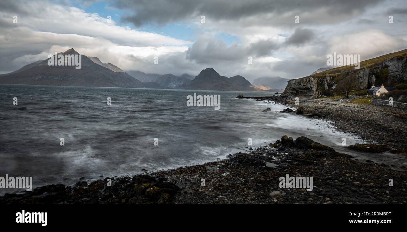 A lake winding through a field underneath a stormy sky with mountains ...