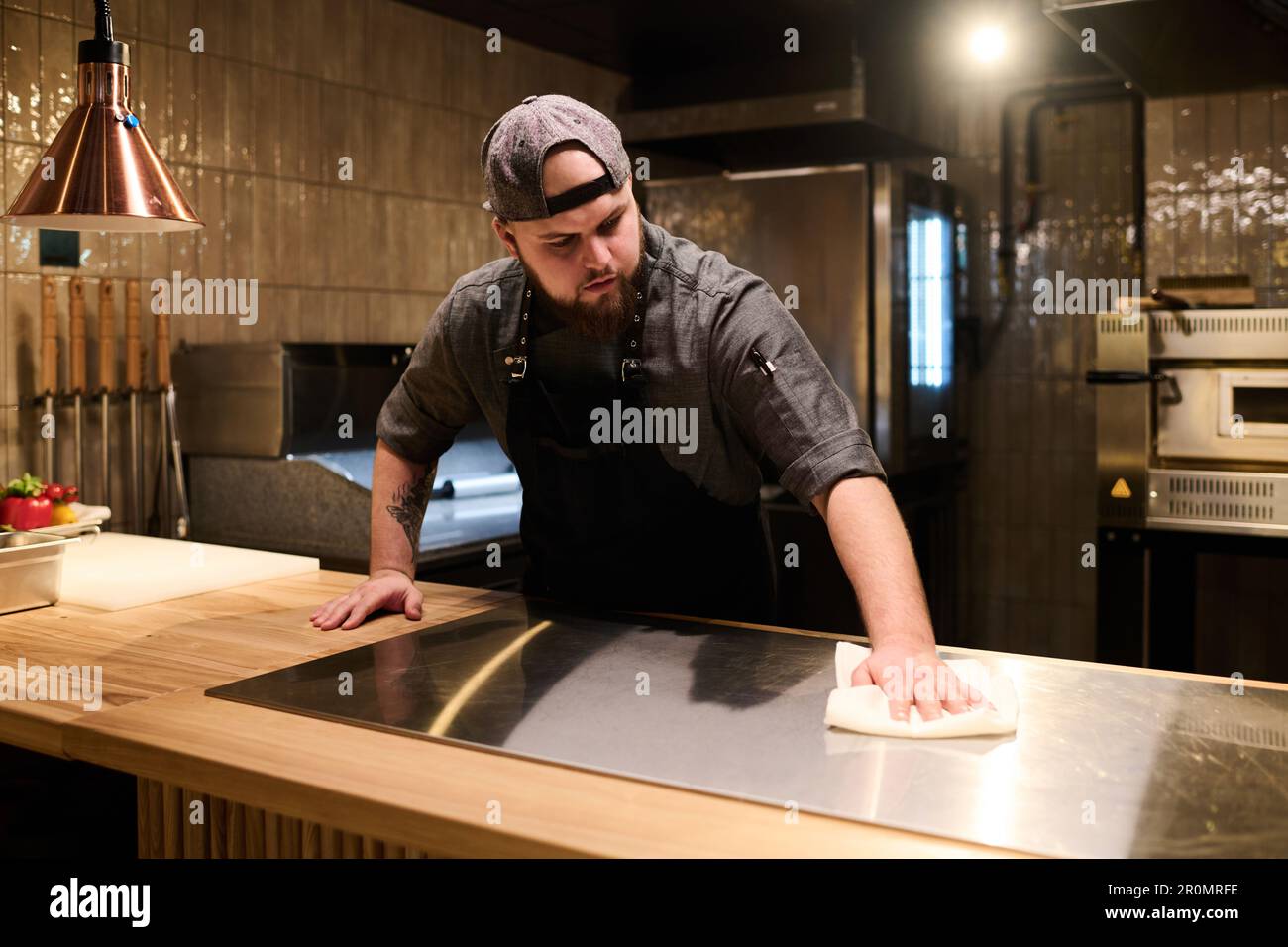 Young male chef in uniform wiping stove surface with clean duster while ...
