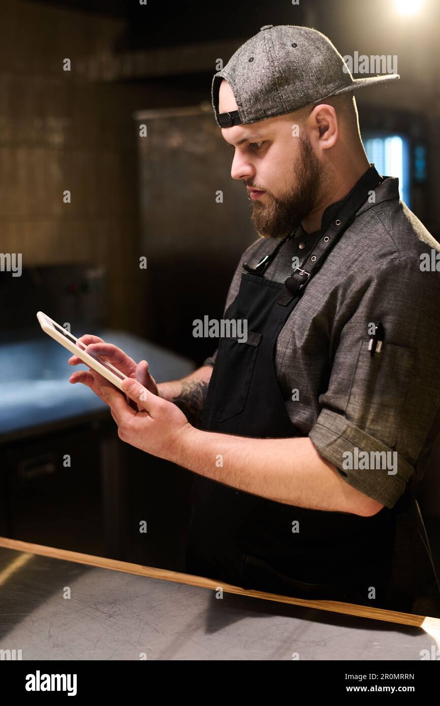 Side view of young serious cook in uniform scrolling through online ...
