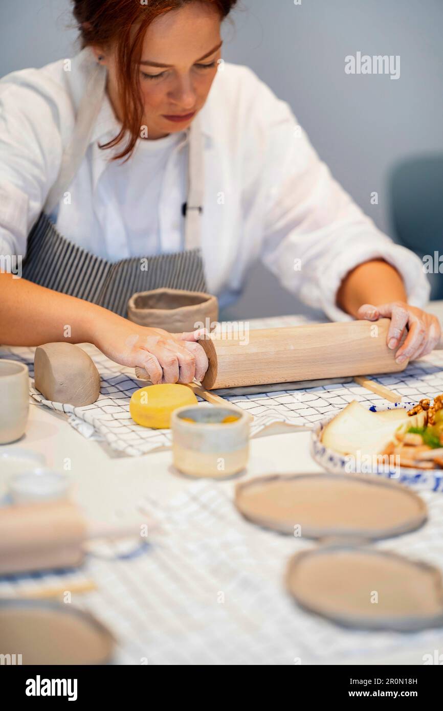 Young female artisan in apron sitting at table and rolling clay slab ...