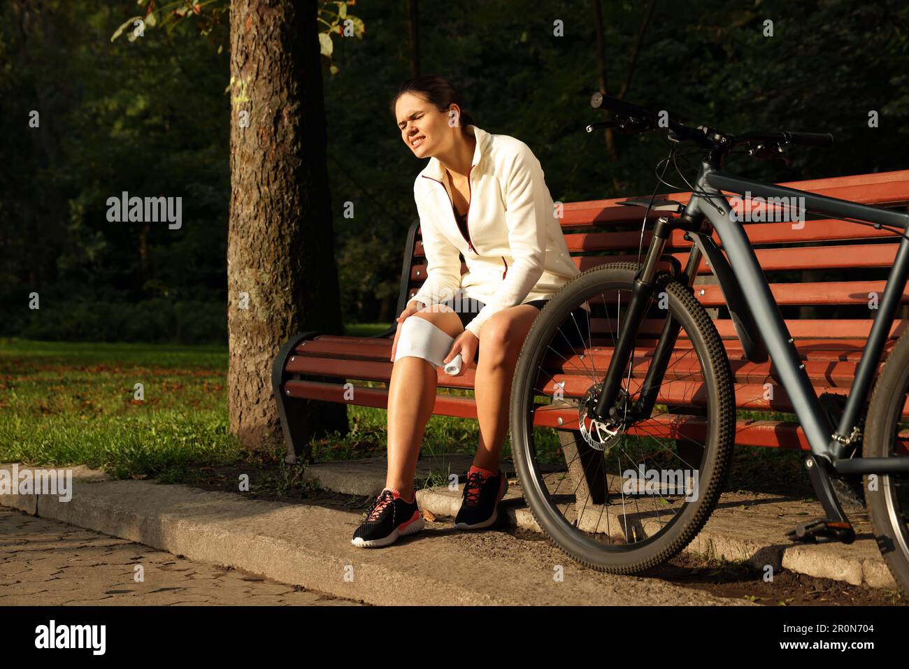 Young woman applying bandage onto her knee on wooden bench outdoors ...