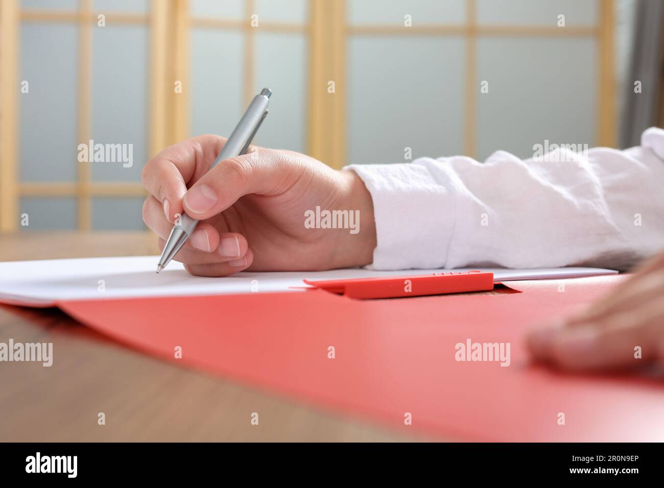 Woman writing on sheet of paper in red folder at wooden table in office ...