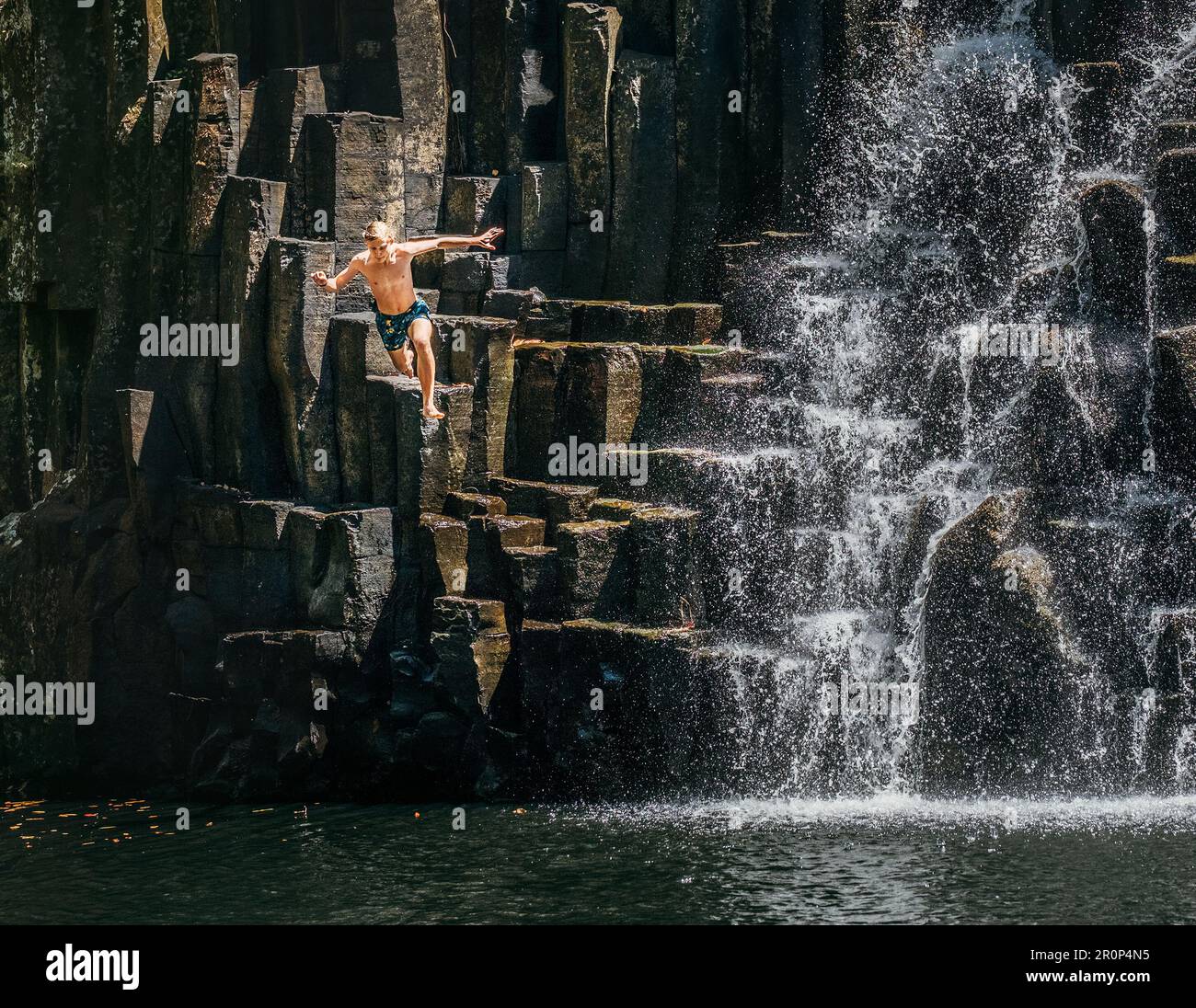 Teenage boy jumping in waterfall lake. Falling water streams flow on ...
