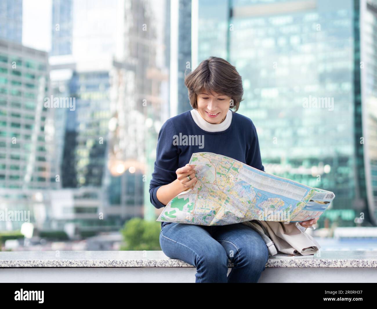 Smiling woman is reading paper map sitting on background of buildings ...