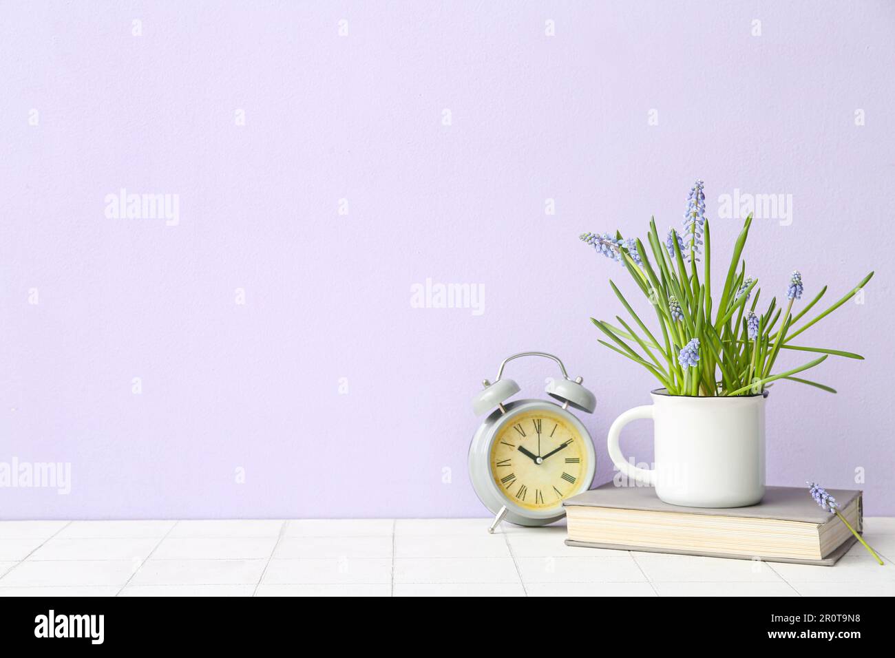 Cup with beautiful Muscari flowers, alarm clock and book on tiled table ...