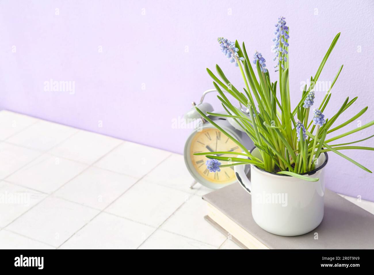 Cup with beautiful Muscari flowers, alarm clock and book on tiled table ...