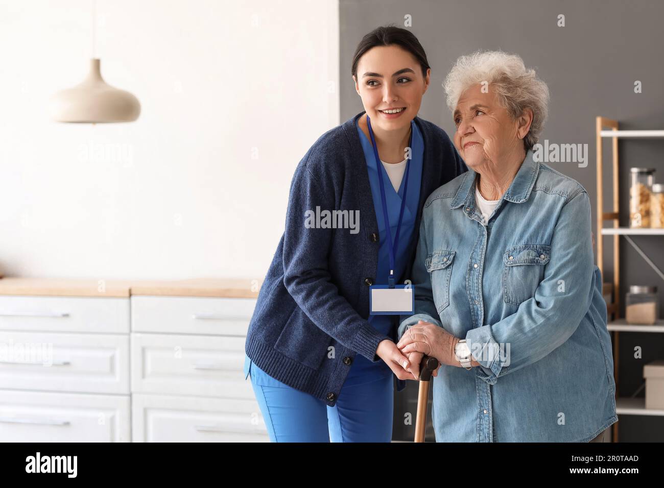 Young caregiver helping senior woman with walking stick in kitchen ...