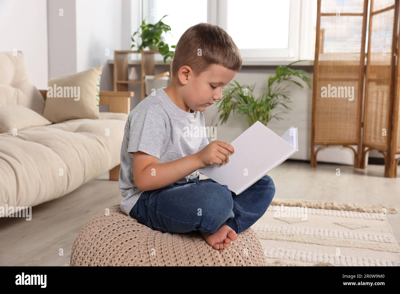 Boy with poor posture reading book on beige pouf in living room ...