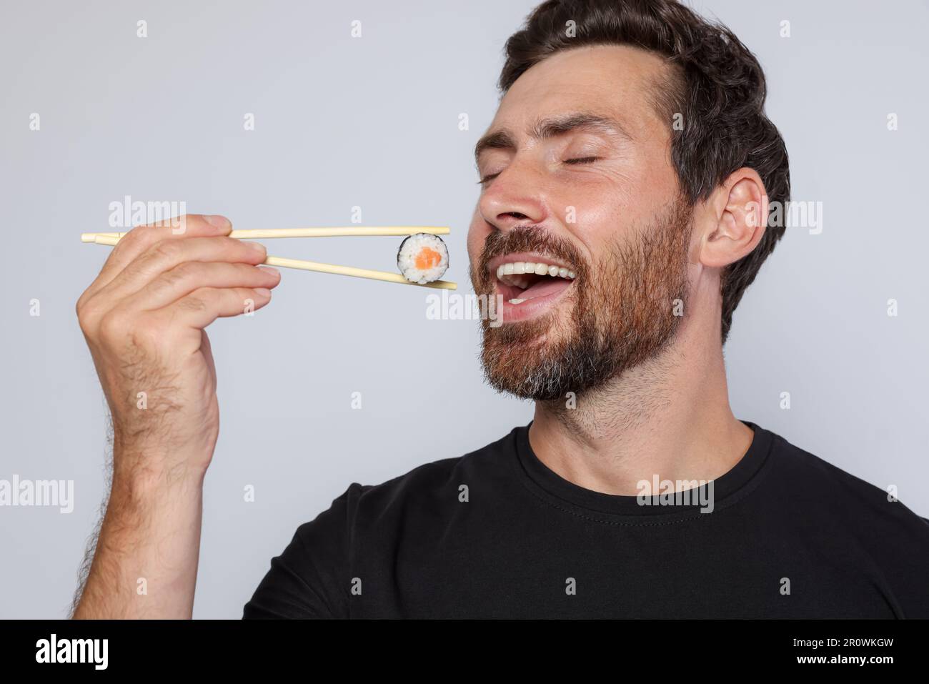 Happy man eating sushi roll with chopsticks on light grey background ...