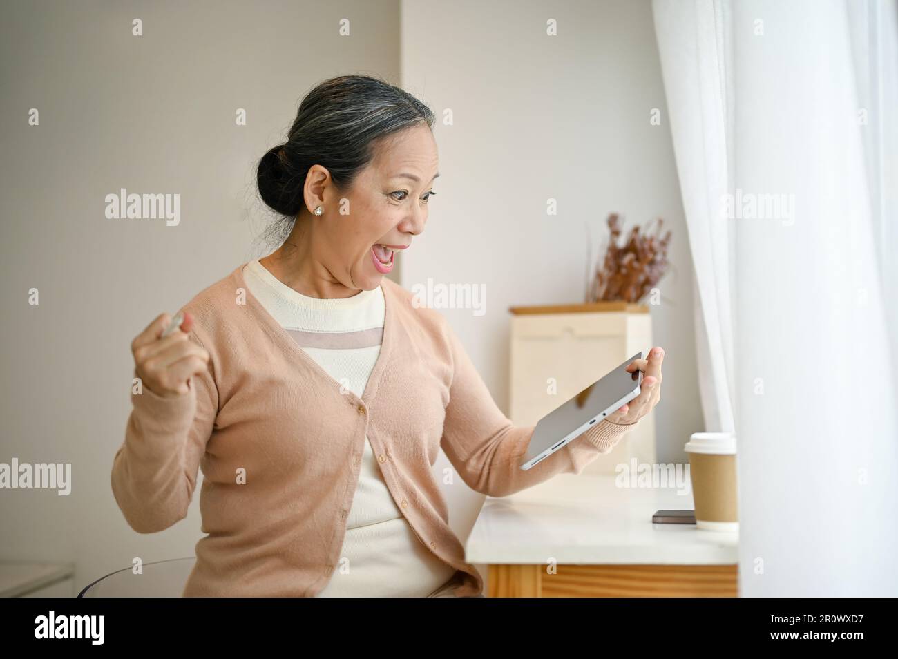 Excited and surprised senior Asian woman sits near the window, looking ...