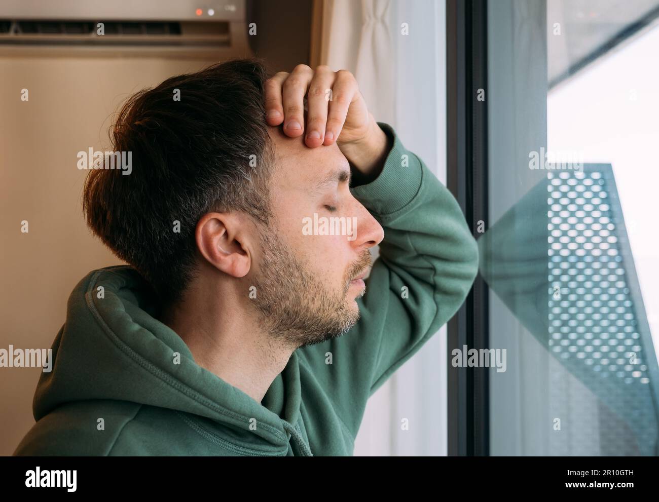 Sad middle age man stands alone at window with closed eyes, holding ...