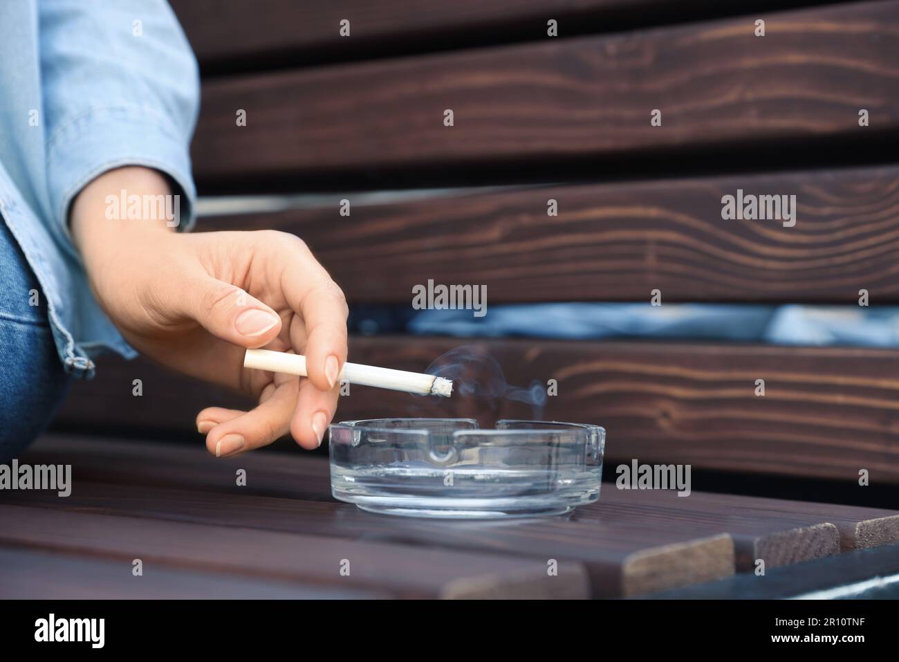 Woman holding cigarette over glass ashtray on bench outdoors, closeup ...