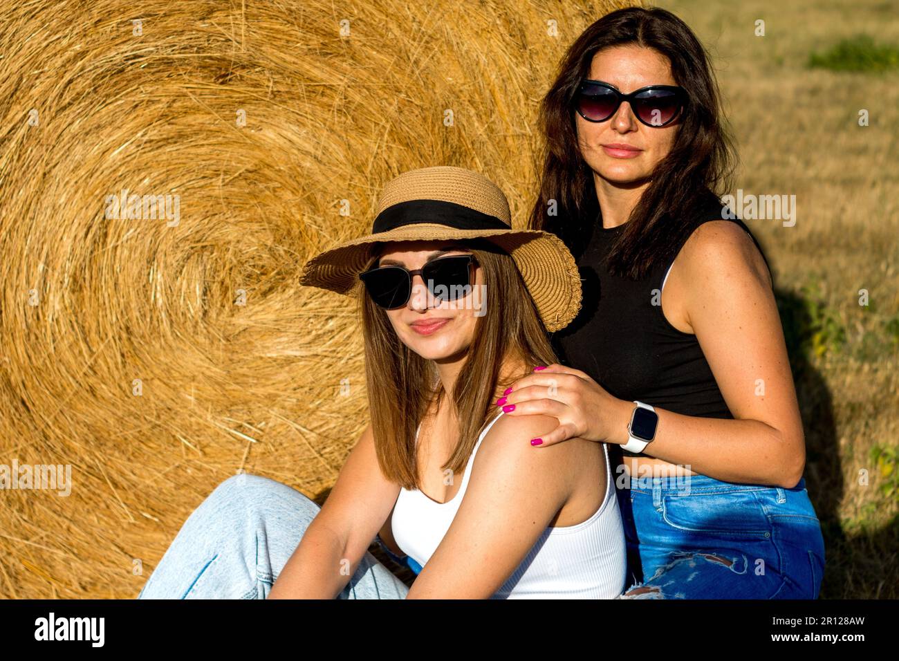 Two Caucasian women, smiling, sitting side by side on a large hay bale ...