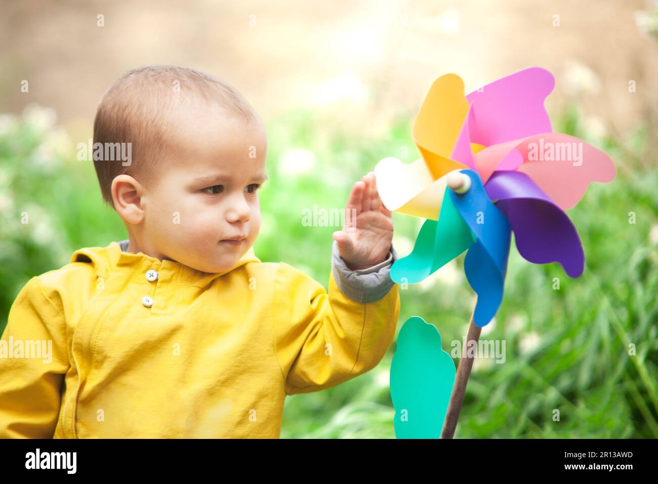 A child with a yellow coat plays with a colorful pinwheel toy in a lush ...