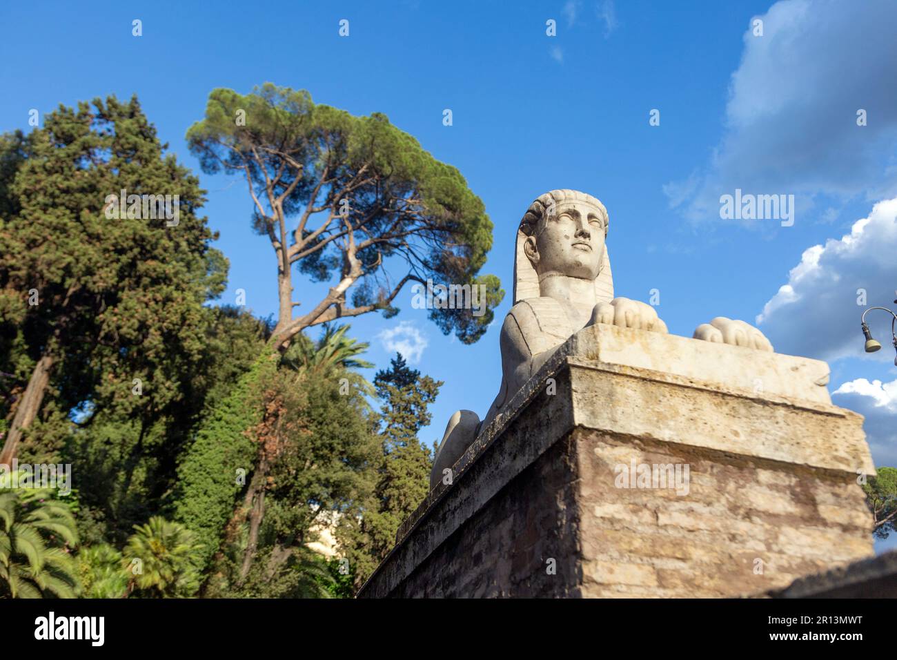 A statue of an ancient lion-sphinx stands in front of a pine tree in ...
