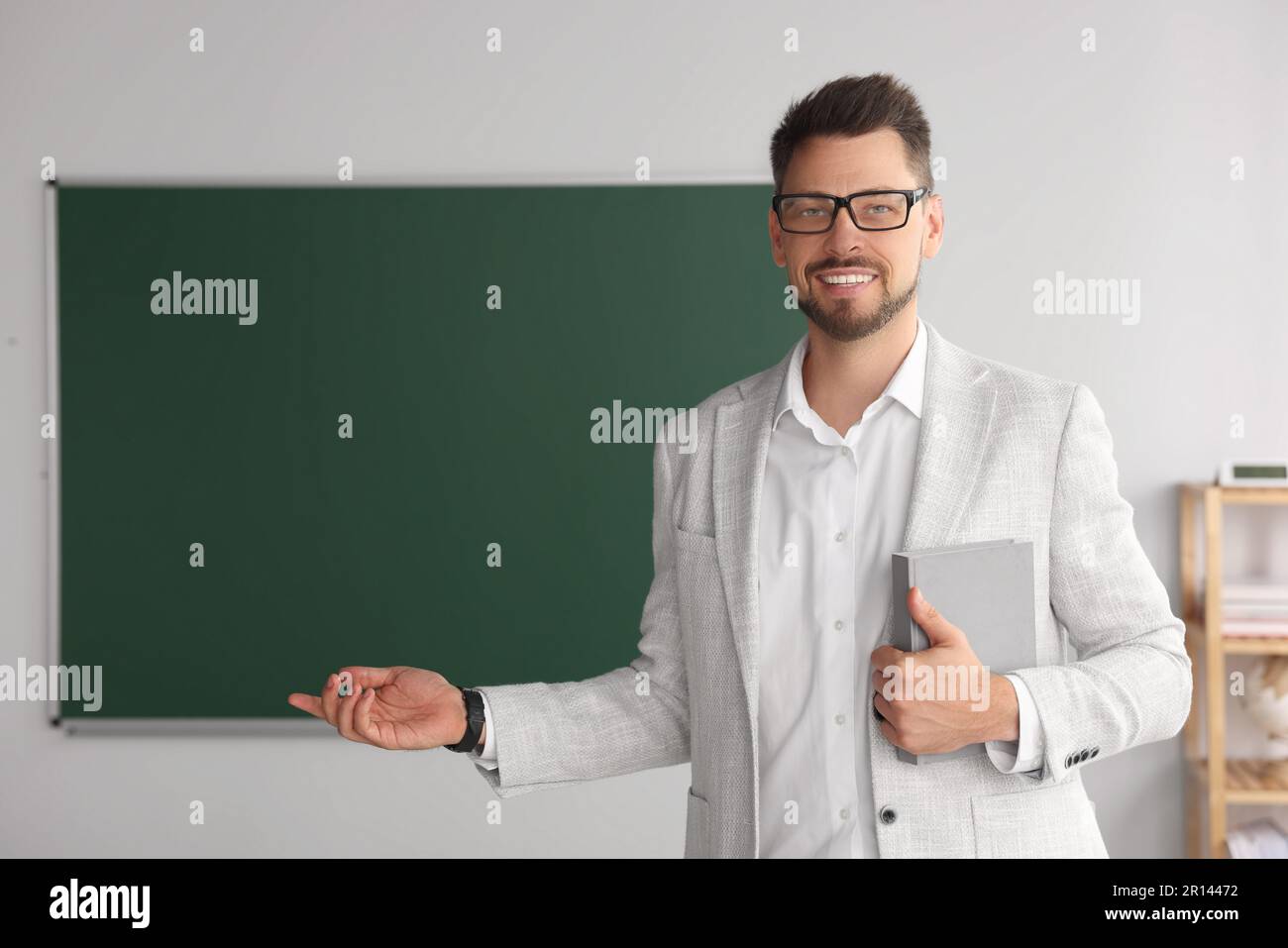 Happy teacher with book explaining something at blackboard in classroom ...