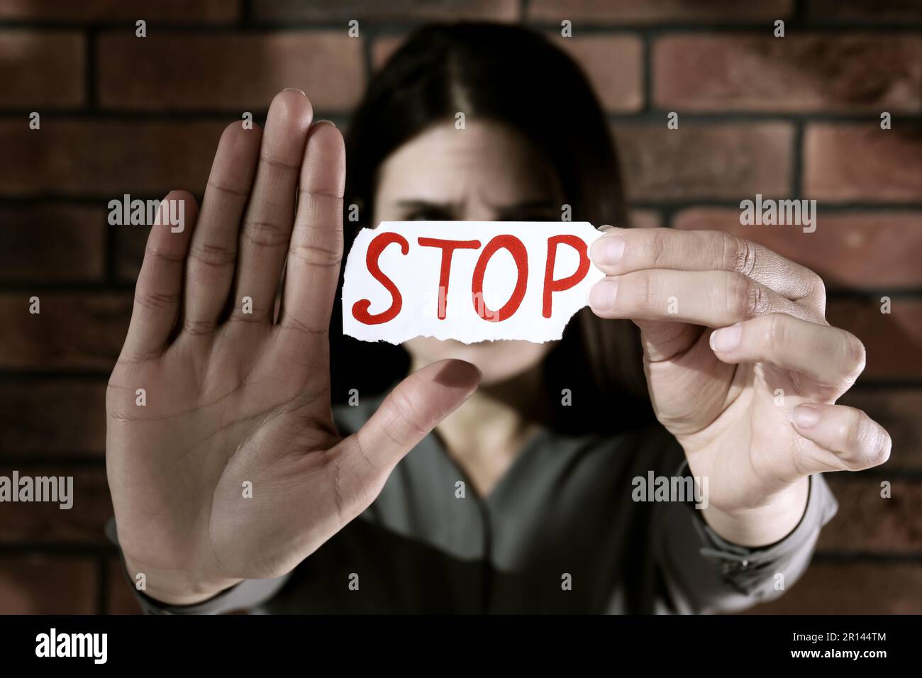 Domestic violence concept. Woman holding paper with word Stop near brick wall, focus on hands ...