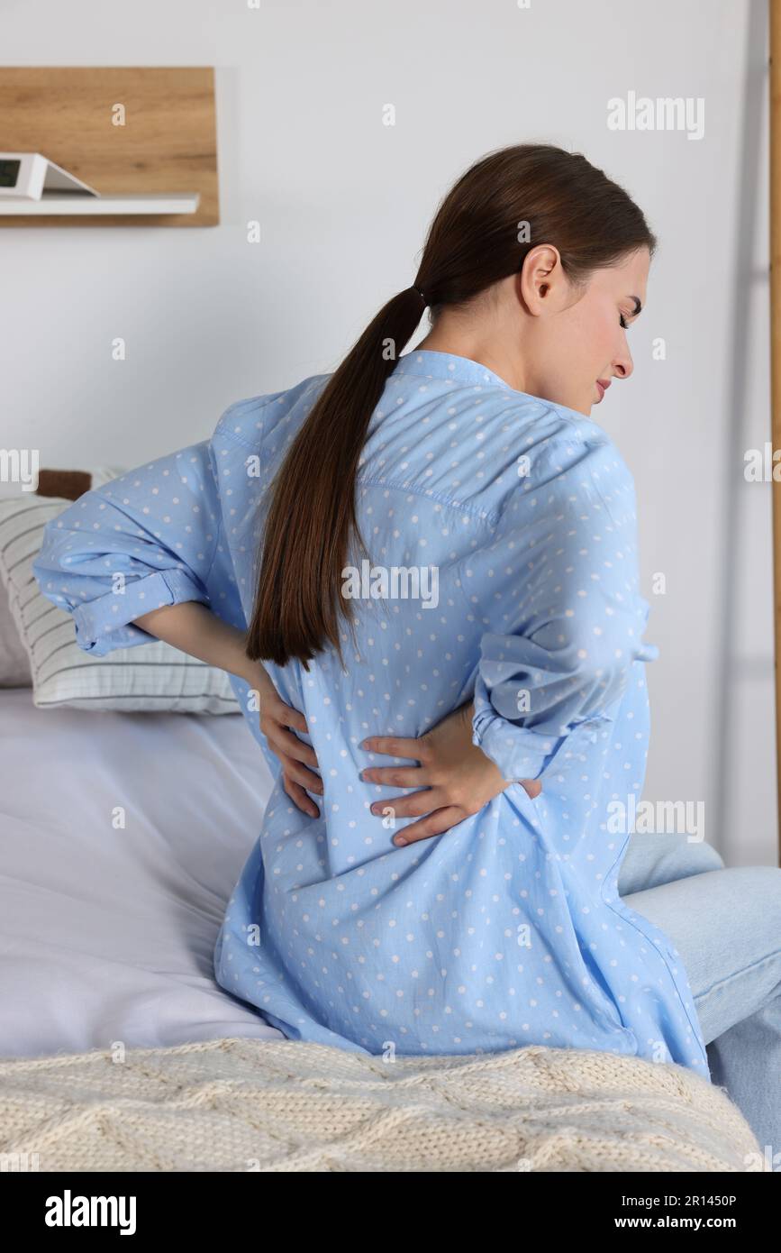 Woman suffering from back pain while sitting on bed at home. Symptom of ...