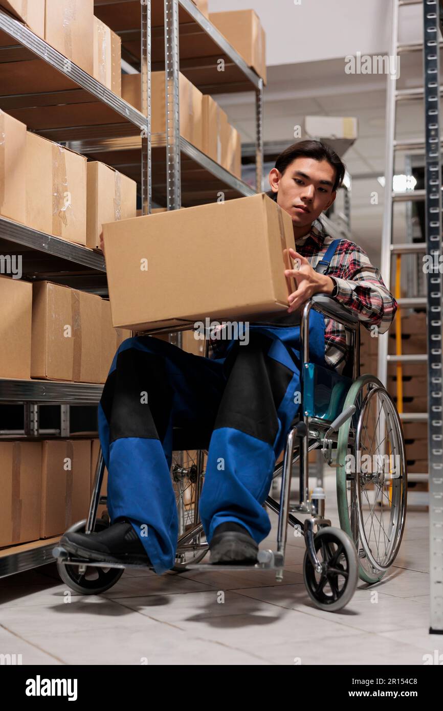 Asian man wheelchair user carrying parcel in postal warehouse aisle ...