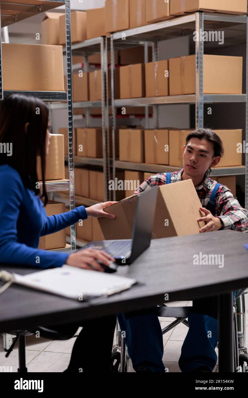 Warehouse manager taking parcel from worker in wheelchair to create invoice on laptop. Post ...