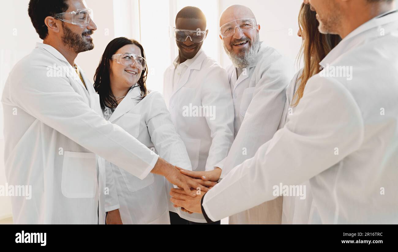 A diverse team of three men and three women, wearing lab coats and safety glasses, join hands in ...