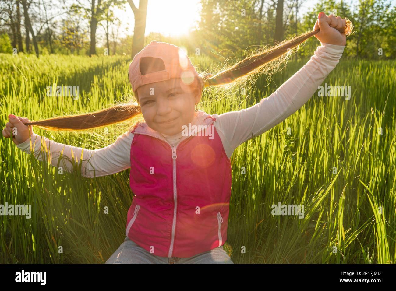 A cheerful girl sits in the thick green grass, holding her pigtails ...
