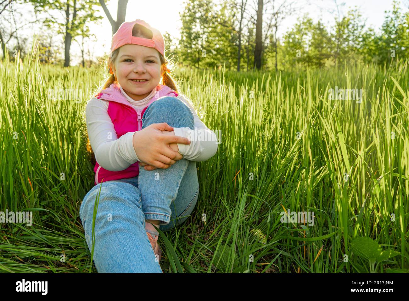 A cheerful girl sits in the thick green grass, clasping one leg with ...