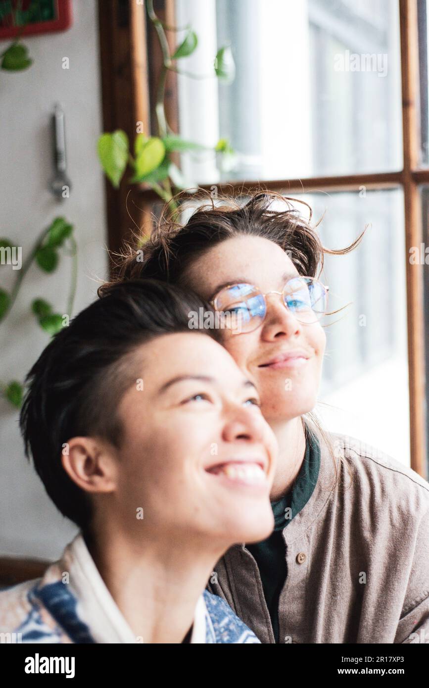 cute queer female couple smile as they cuddle by window at home Stock ...